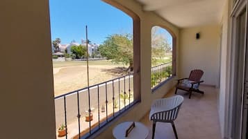Uma varanda com duas cadeiras e uma mesa pequena, com vista para uma área arenosa e edifícios ao longe.
