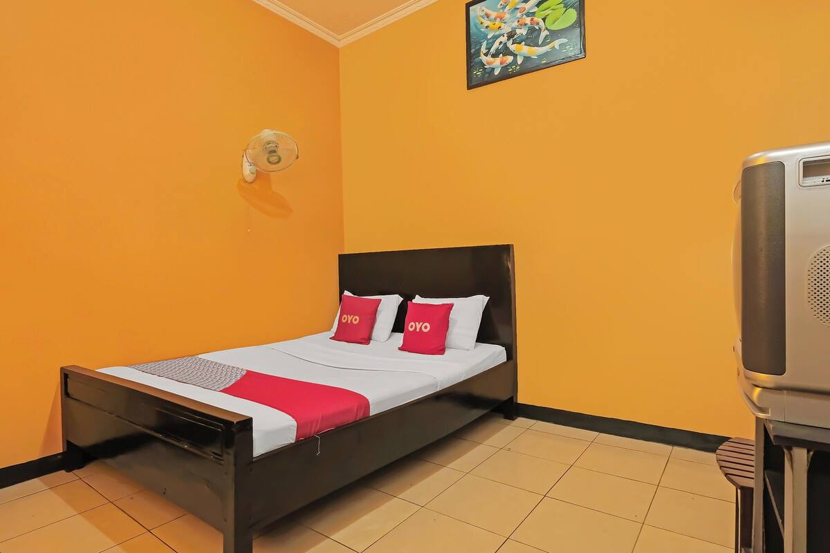 Uma cama de solteiro com estrutura preta, roupa de cama branca e almofadas vermelhas. Há uma pequena mesa com uma televisão no lado direito do quarto. As paredes são laranja e há um quadro na parede acima da cama.