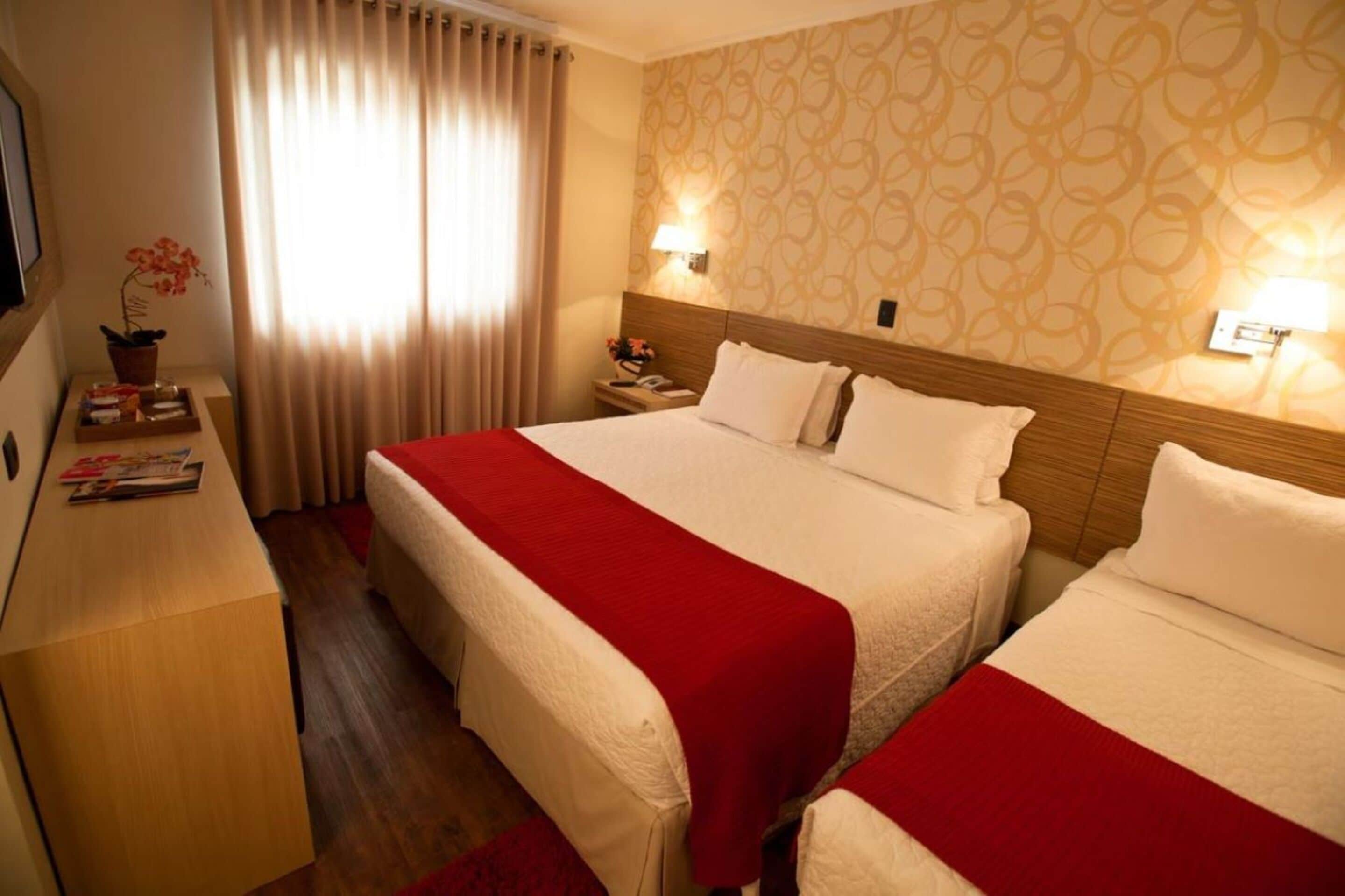 Quarto de hotel com duas camas, uma cabeceira de madeira, uma escrivaninha com luminária, uma planta em vaso e uma janela com cortinas.