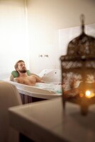 Uma pessoa relaxando em uma banheira com um apoio de cabeça verde, uma parede branca com um porta-toalhas e um objeto decorativo no balcão.