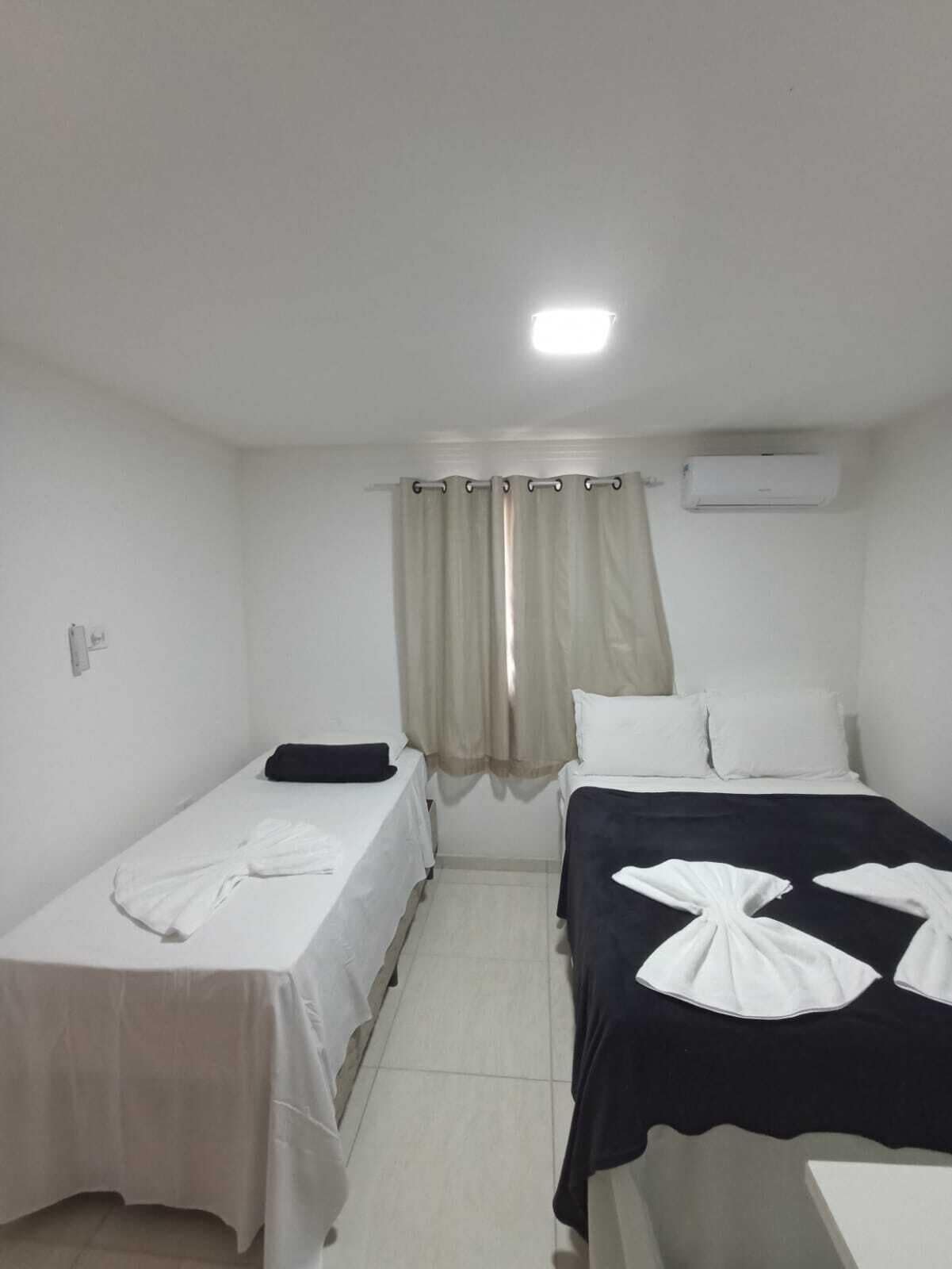 Quarto pequeno e limpo com duas camas de solteiro, janela com cortinas, aparelho de ar condicionado e parede branca.