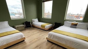 Quarto com três camas, cada uma com estrutura de madeira e roupa de cama branca, dispostas em formato de U. Janelas amplas oferecem vista para o exterior.