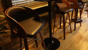 Uma área de bar com cadeiras e mesas de madeira, paredes de tijolo e uma janela com persianas.