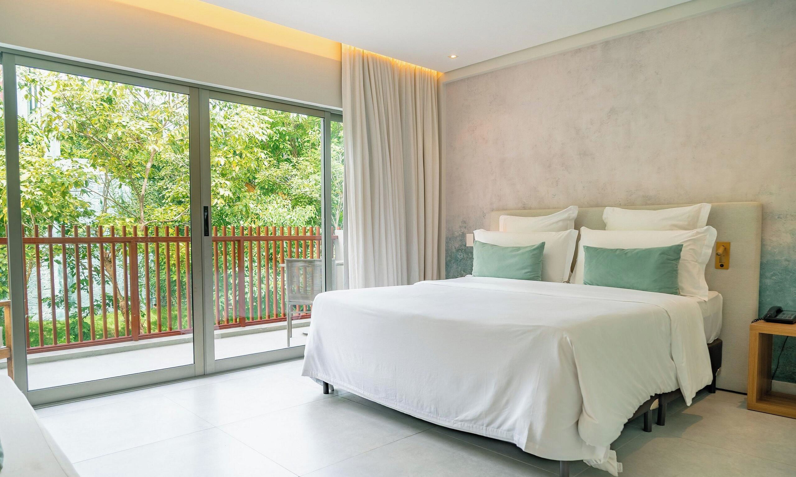 Quarto moderno com uma cama grande, mesa de cabeceira e vista para o verde através de uma porta de vidro deslizante.