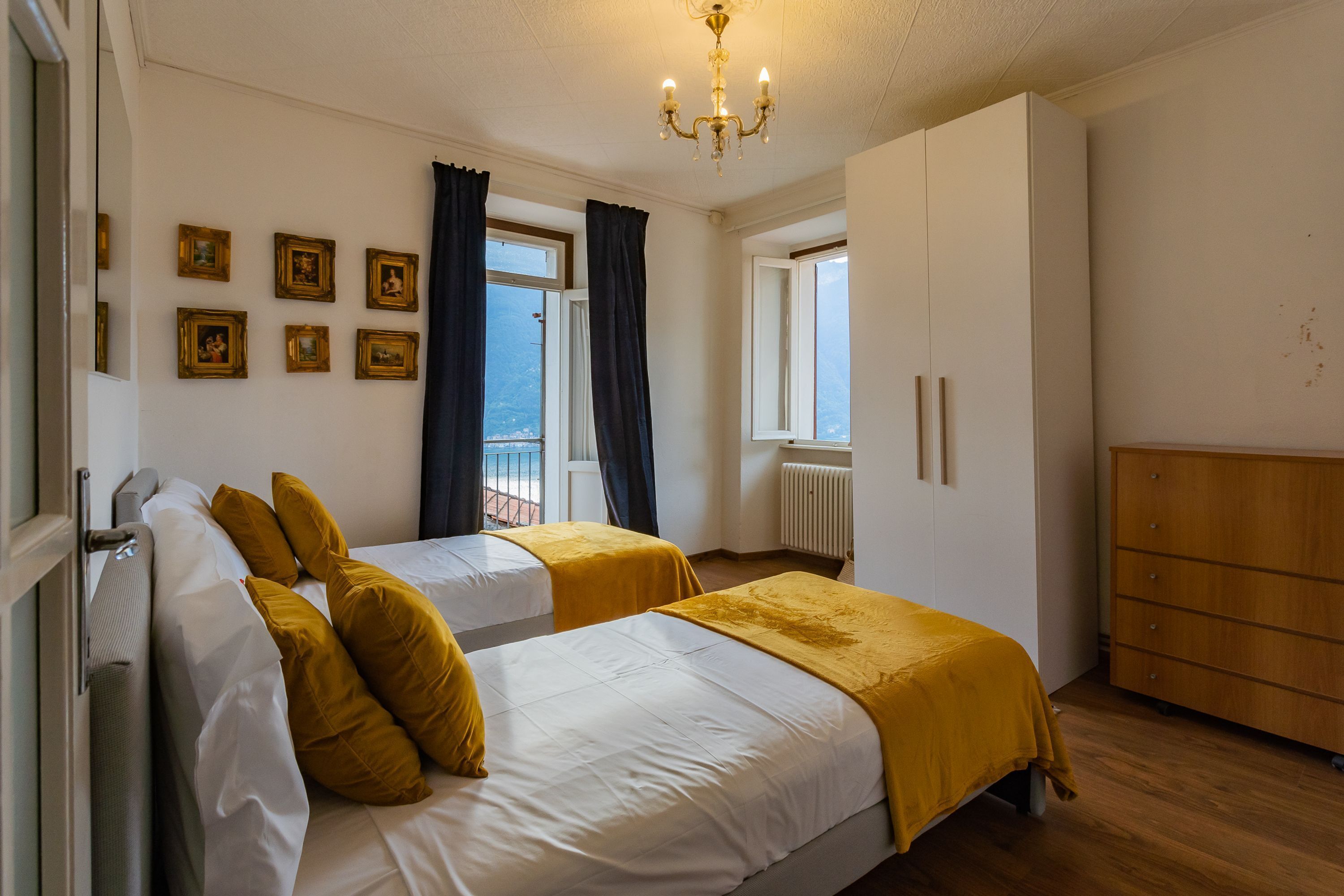 Quarto com duas camas, cômoda de madeira, lustre e janela com cortinas.