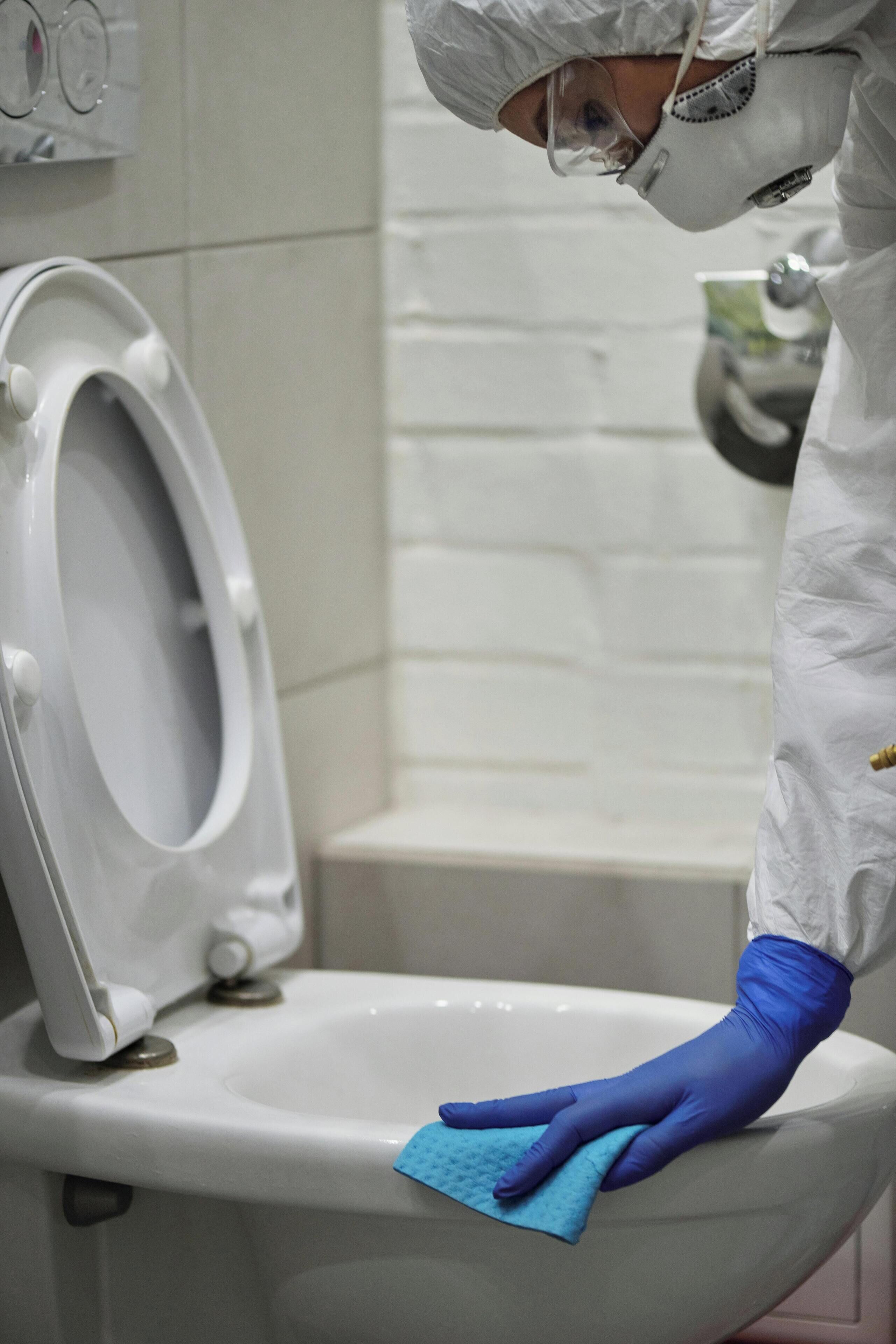 Uma pessoa vestindo um traje de proteção branco limpando um vaso sanitário com um pano azul.