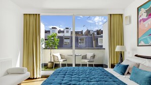 Quarto com uma cama grande, um sofá branco, uma mesa pequena e uma cadeira. Há uma janela com cortinas, vista de prédios lá fora e um quadro na parede.