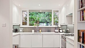 Cozinha moderna com armários brancos, uma janela grande com vista para vegetação, fogão a gás e uma estante com livros.
