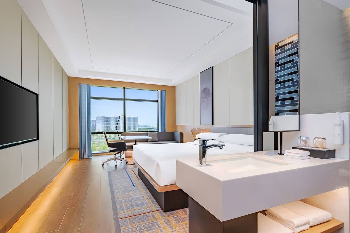 Quarto de hotel moderno com uma cama grande, banheiro com pia espaçosa e televisão fixada na parede.
