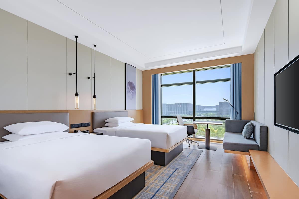 Quarto de hotel moderno com duas camas, um sofá, uma TV e uma janela grande com vista para a cidade.