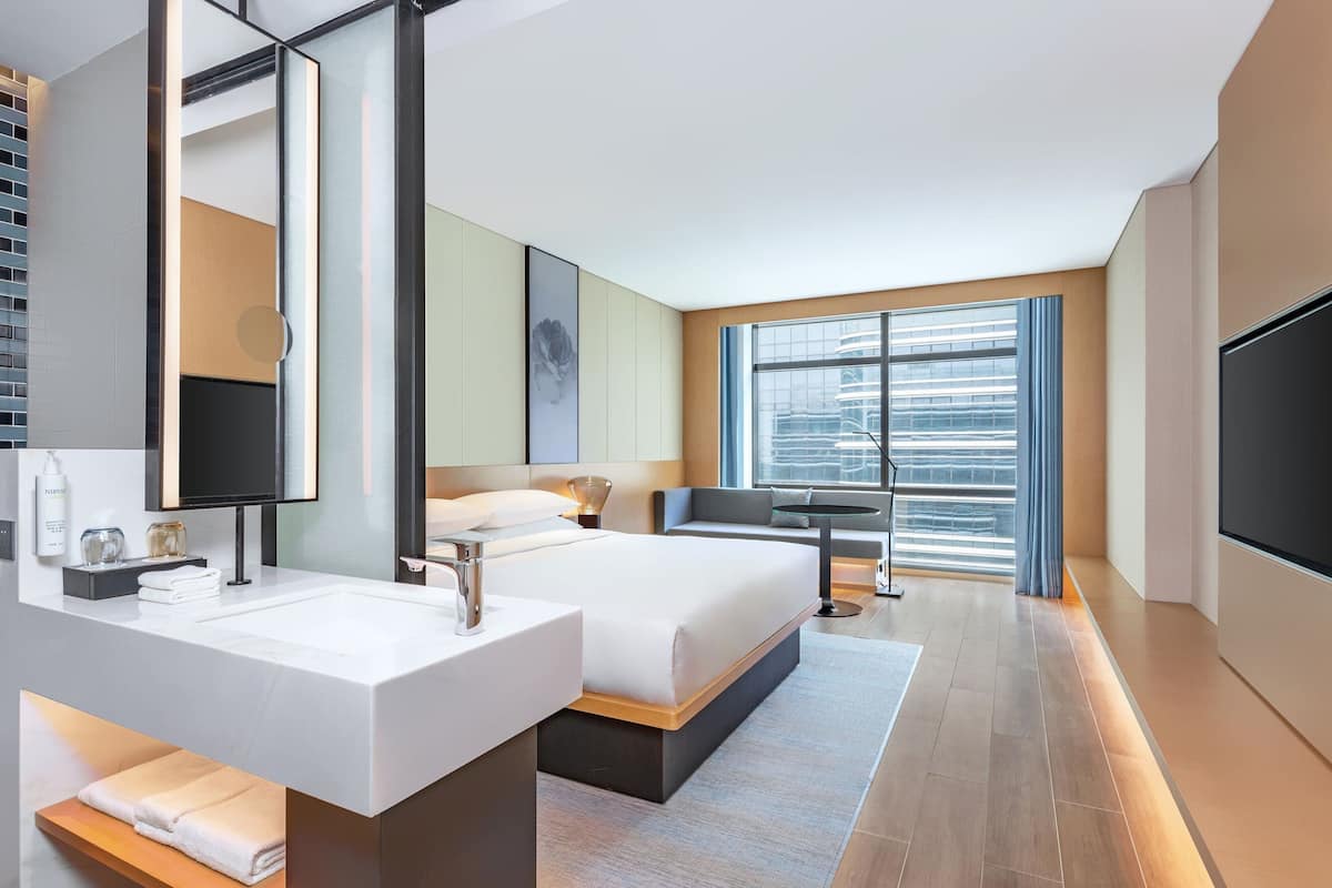 Quarto de hotel moderno com uma cama grande, duas mesas de cabeceira, um espelho amplo, uma televisão e uma janela com vista para a cidade.
