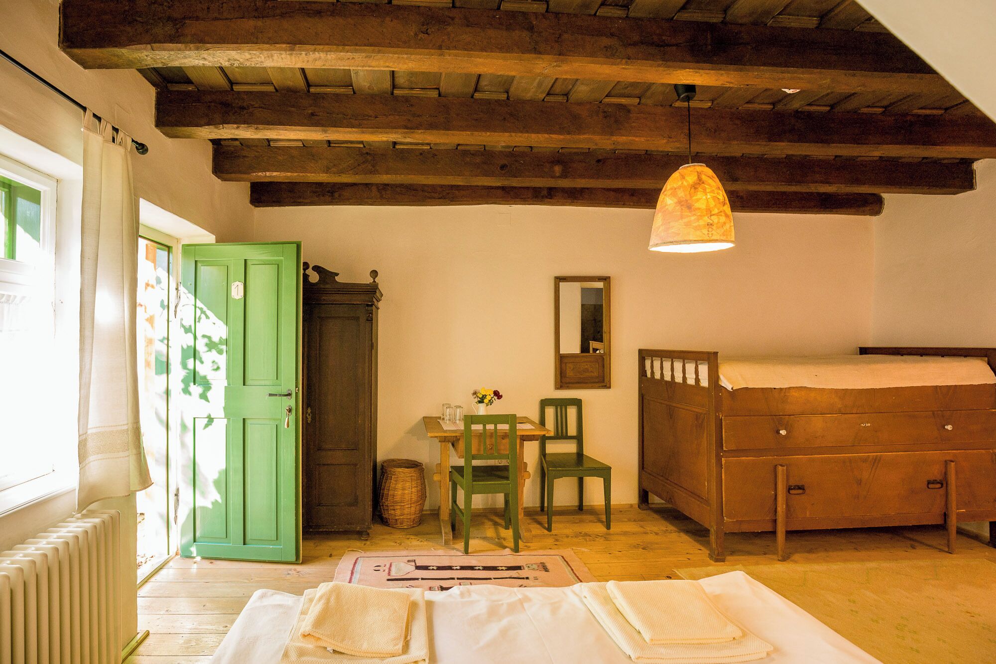 Quarto com teto de madeira, porta verde, cômoda de madeira, mesa com cadeiras e uma cama.