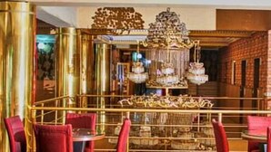 Um hall luxuoso com um lustre imponente, poltronas estofadas em vermelho e um corrimão dourado.