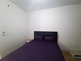 Quarto pequeno com uma cama de solteiro, coberta por um edredom roxo, paredes brancas e assoalho de madeira.