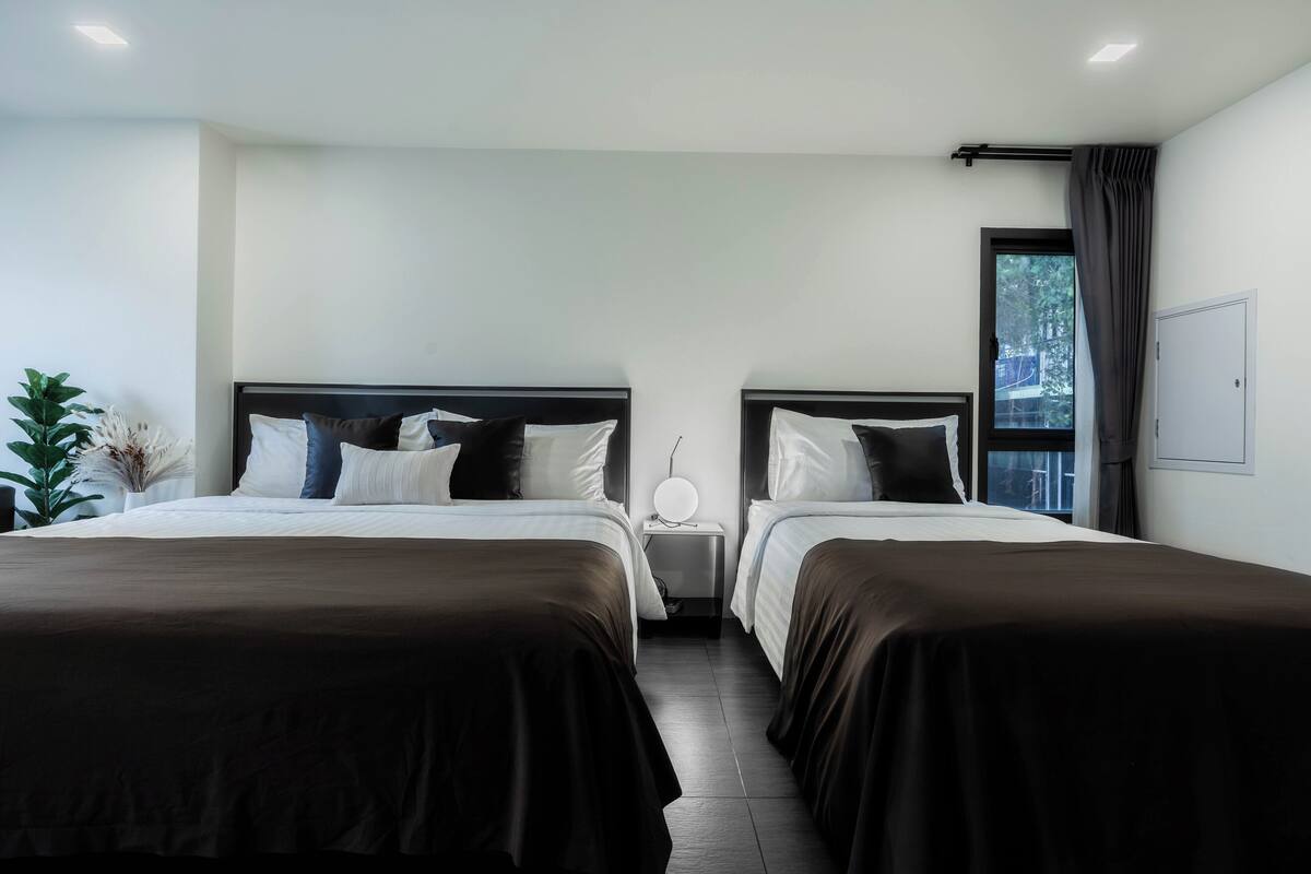 Quarto de hotel com duas camas, uma mesinha de cabeceira com abajur e uma janela com cortinas.