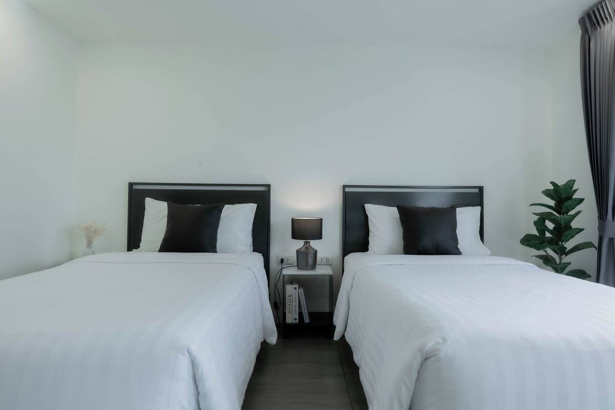 Quarto de hotel com duas camas de solteiro, uma mesa de cabeceira com abajur, uma pequena planta e uma mesinha de cabeceira com livros.