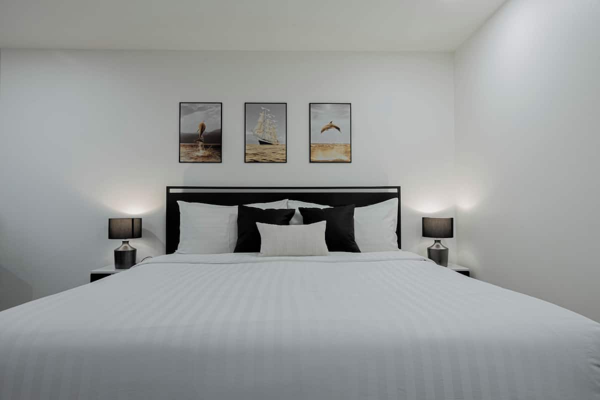 Cama bem arrumada com roupa de cama branca, com duas mesas de cabeceira e luminárias, e três quadros na parede acima.