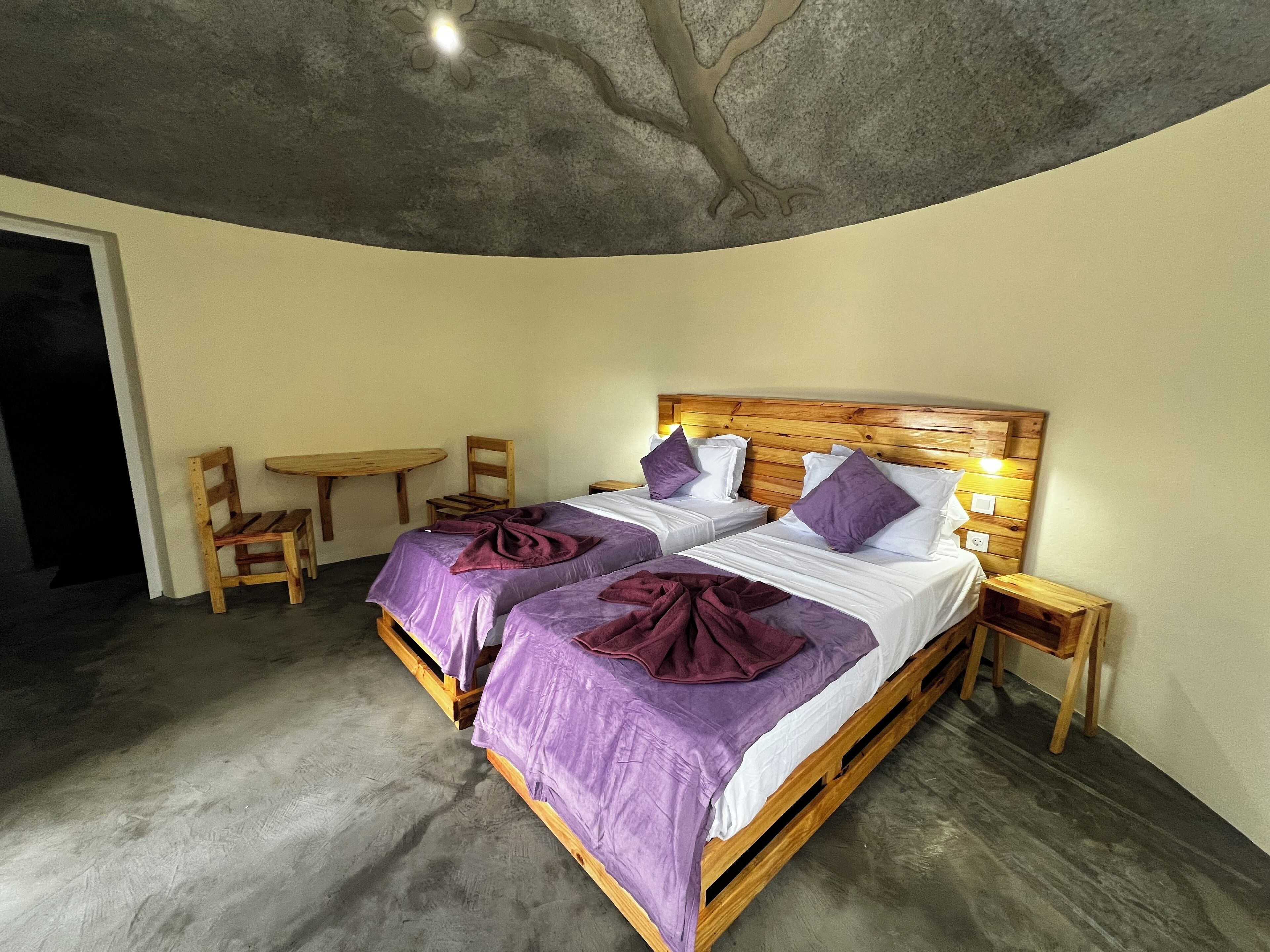 Uma cama de casal com cabeceira de madeira e duas mesas de cabeceira, uma pequena mesa de madeira e uma cadeira em um quarto com teto de concreto.