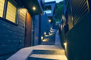 Uma escada estreita e bem iluminada conduzindo a um segundo andar, ladeada por revestimento de madeira e uma parede de concreto.