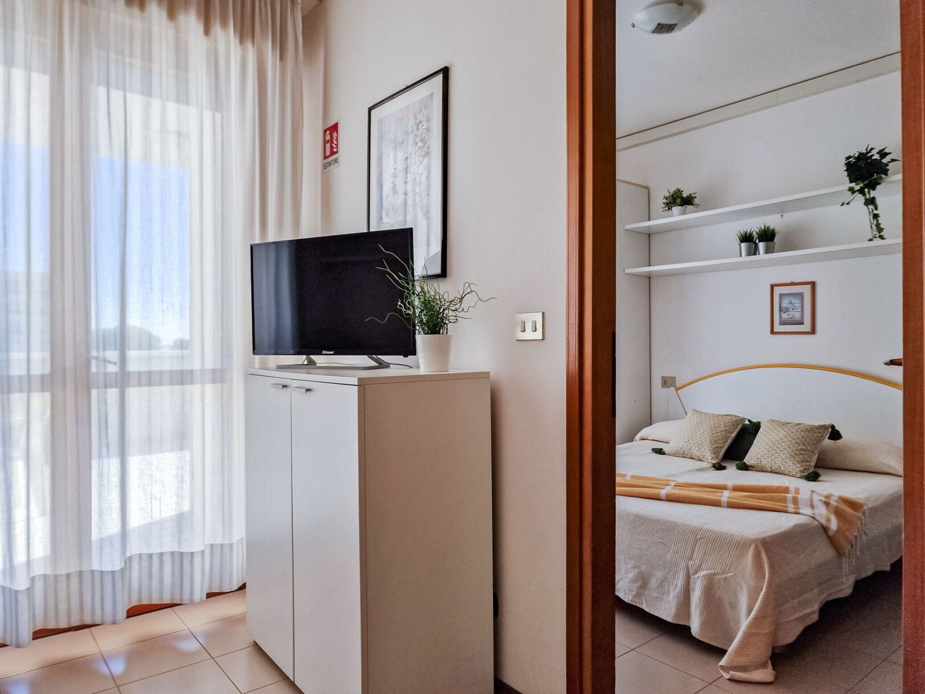 Quarto moderno com cama, televisão em suporte, planta e prateleira com objetos decorativos.