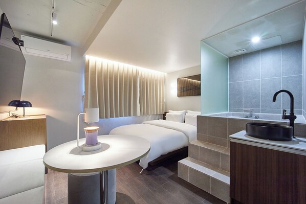 Quarto de hotel pequeno e funcional, com cama, mesinha redonda com abajur, pia embutida e chuveiro.
