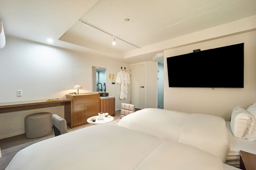 Quarto de hotel moderno com uma cama grande, uma escrivaninha com abajur, toucador com espelho e uma televisão na parede.