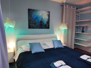 Quarto com cama, travesseiros, um quadro, mesa de cabeceira e prateleira.