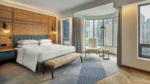 Quarto de hotel moderno com uma cama grande, um banquinho, uma poltrona e vista da cidade através de amplas janelas.