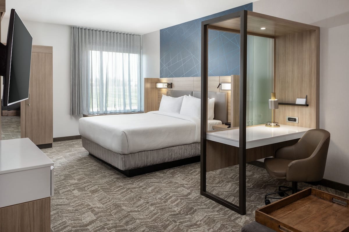 Quarto de hotel com cama, escrivaninha, cadeira, televisão e janela com cortinas.