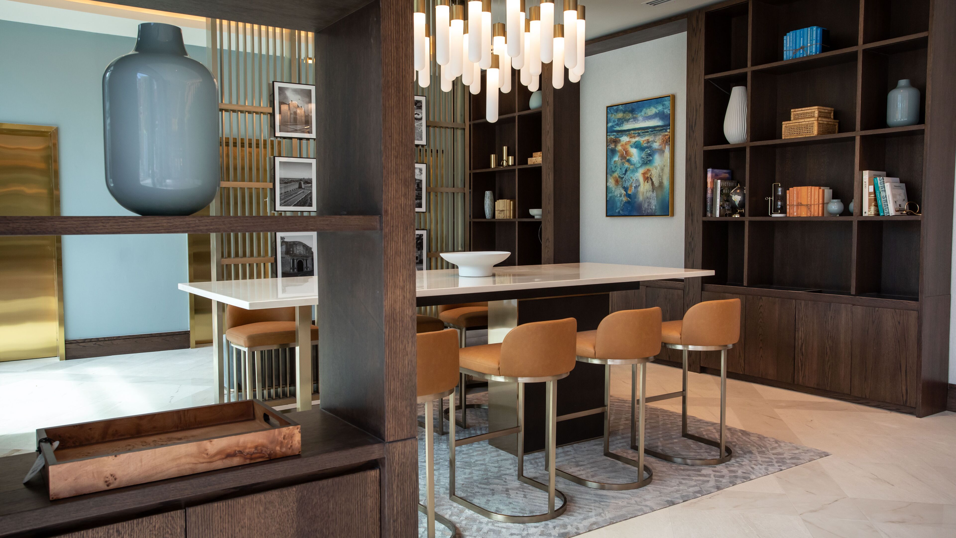 Uma sala de jantar moderna com uma mesa branca e quatro banquetas altas, um vaso grande e uma estante repleta de livros e objetos decorativos.
