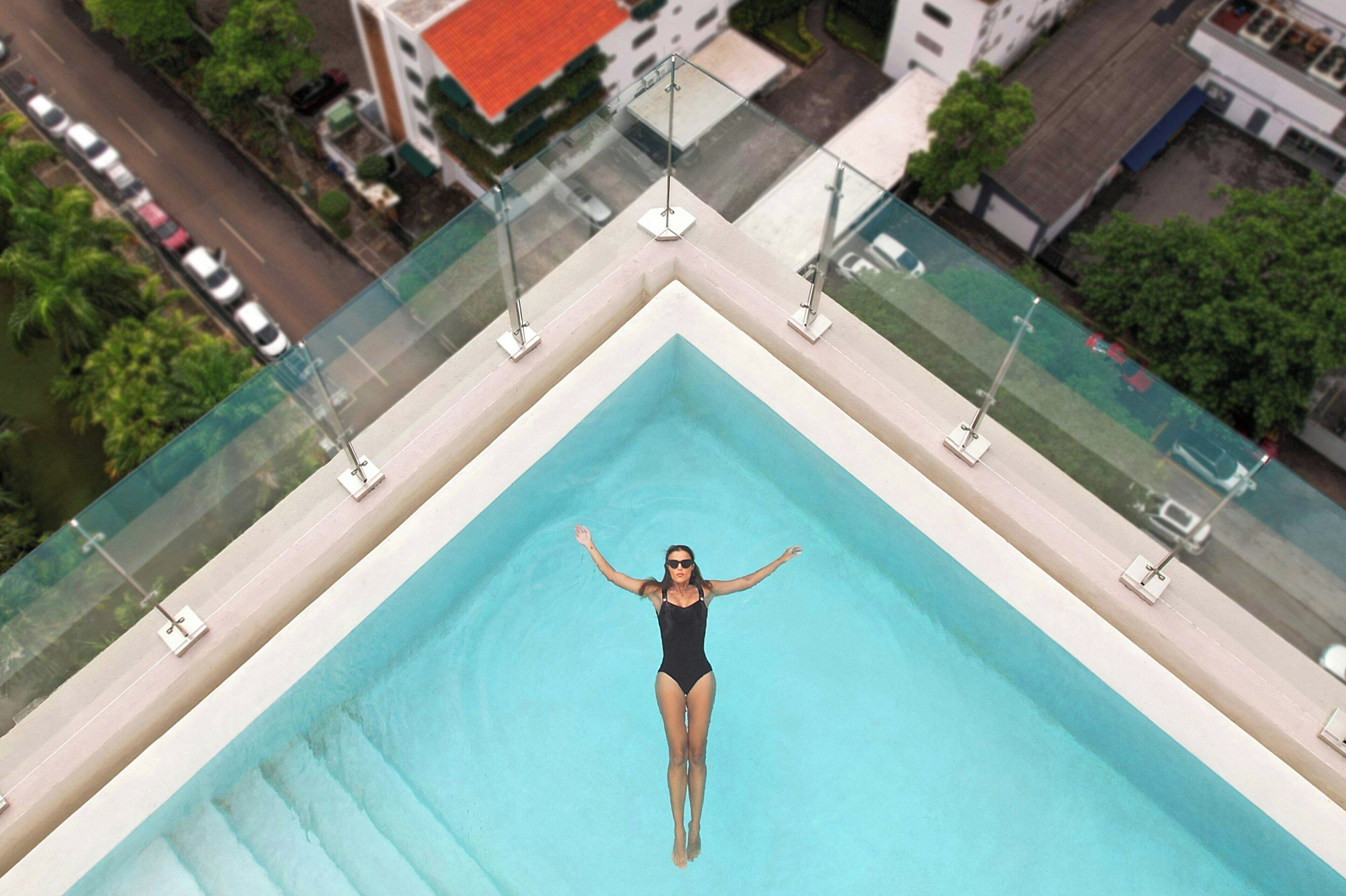 Uma pessoa flutua em uma piscina nos fundos, com grades de vidro e vista para a cidade.