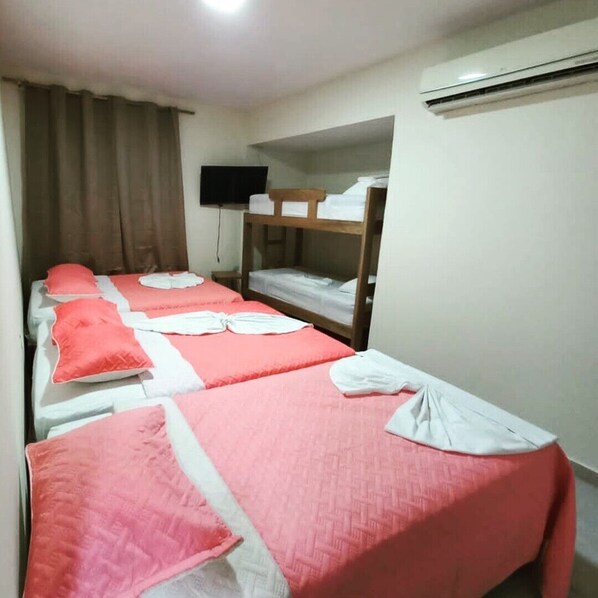 Quarto com duas camas, cada uma com roupa de cama rosa e branca, uma TV de tela plana fixada na parede e um aparelho de ar condicionado sobre as camas.
