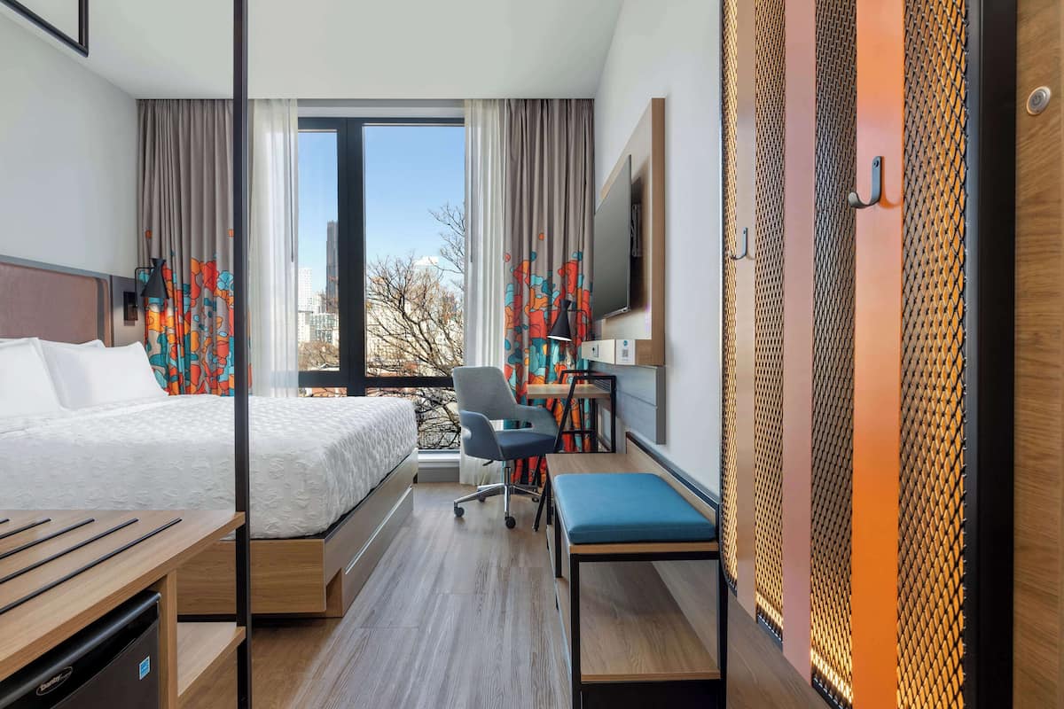 Quarto de hotel com cama, escrivaninha, cadeira e uma janela grande.