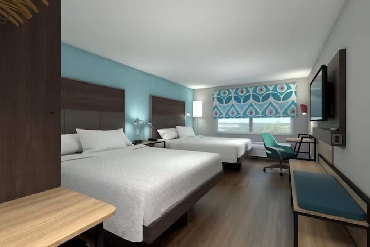 Quarto de hotel com duas camas, uma escrivaninha, uma cadeira, uma televisão e uma janela com cortinas estampadas.