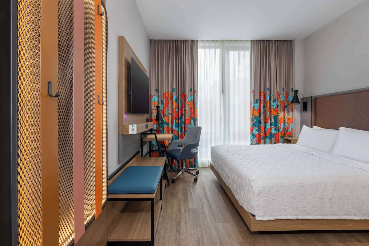 Quarto de hotel com cama, escrivaninha, cadeira e televisão.