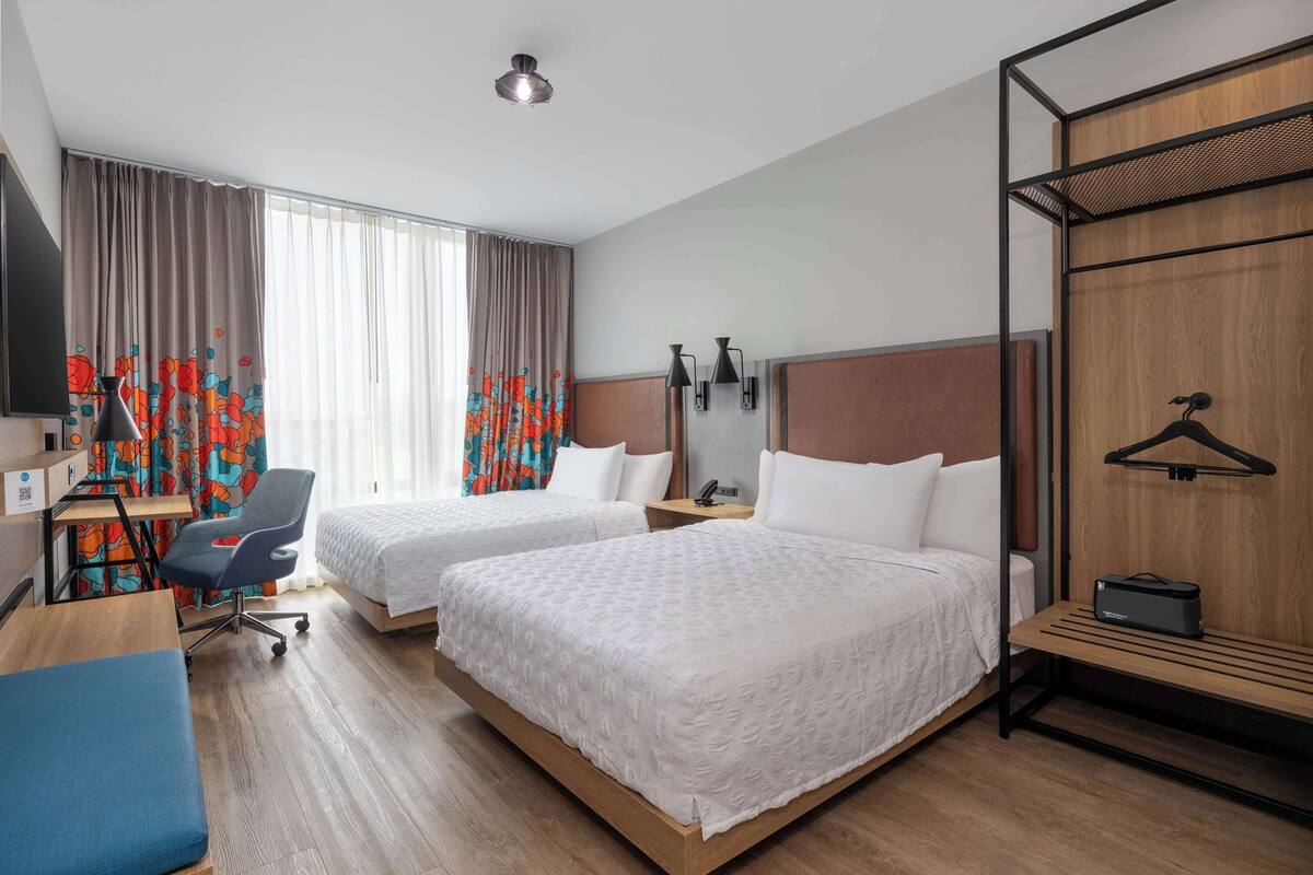 Quarto de hotel com duas camas, uma escrivaninha com cadeira, televisão e guarda-roupa com cabideiro.