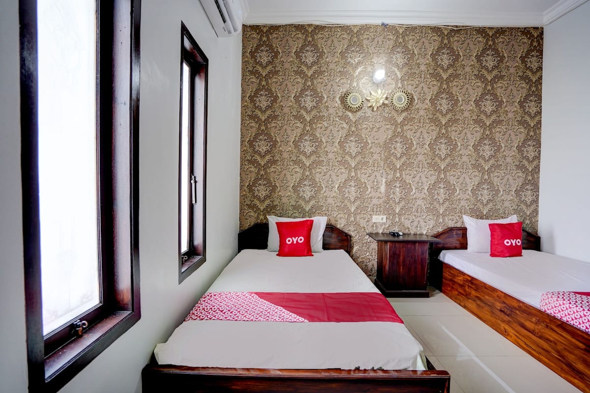 Quarto de hotel com duas camas, cada uma com cabeceira de madeira, papel de parede estampado e travesseiro vermelho com a inscrição 'OYO'.
