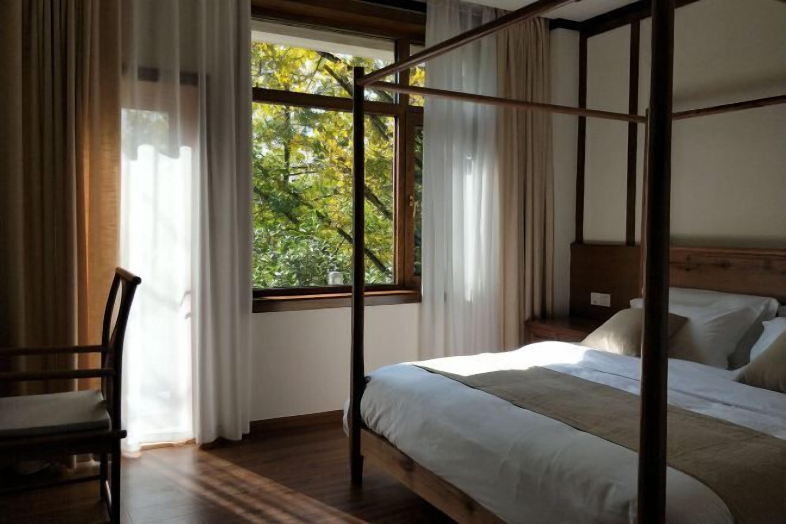 Uma cama de dossel com estrutura de madeira, uma cadeira e uma janela com cortinas.