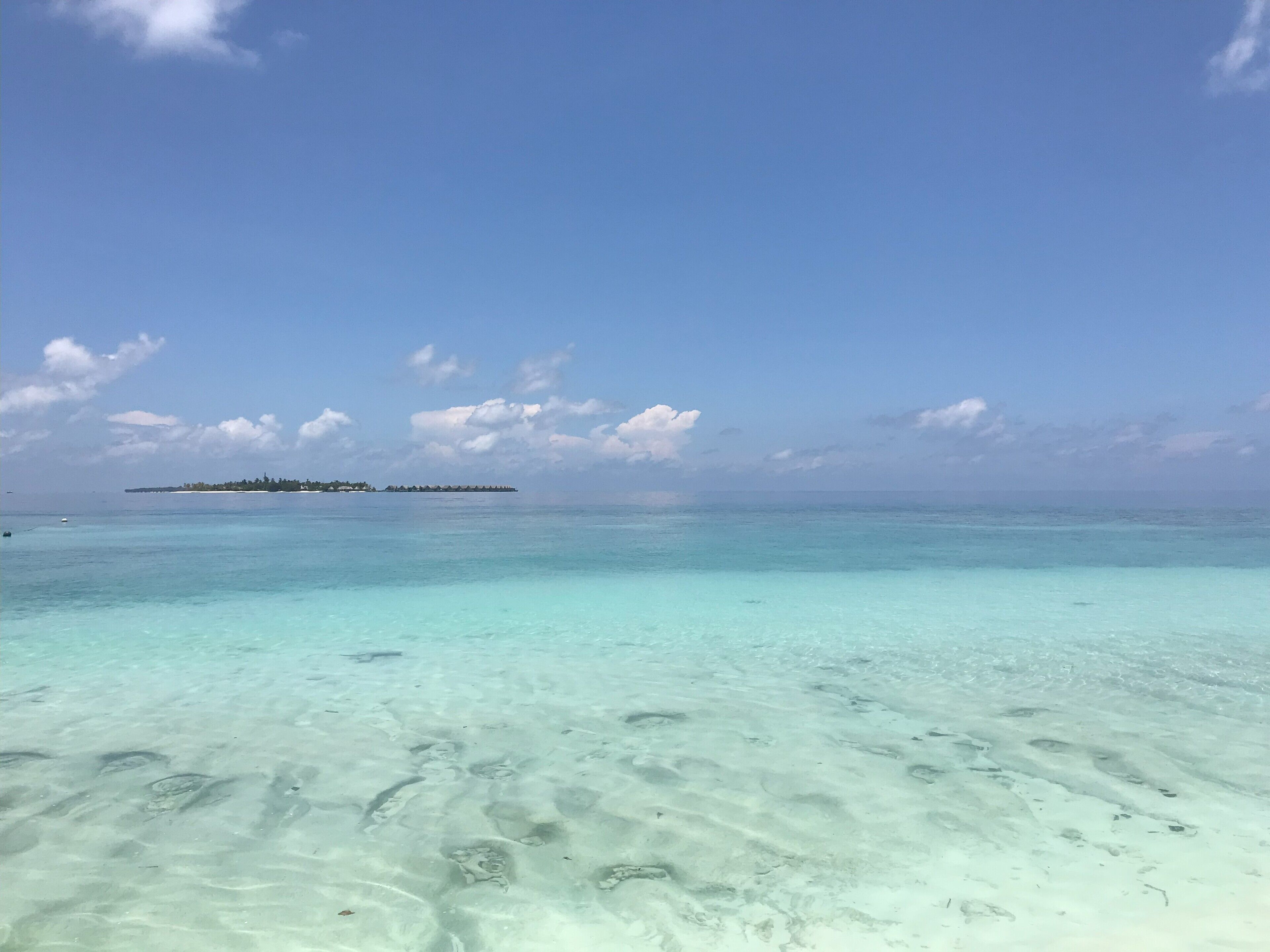 Mar turquesa transparente com uma praia de areia e uma ilha distante com construções.