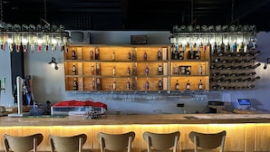 Uma área de bar com prateleiras de madeira exibindo garrafas e copos, um balcão com banquetas e uma televisão sobre as prateleiras.