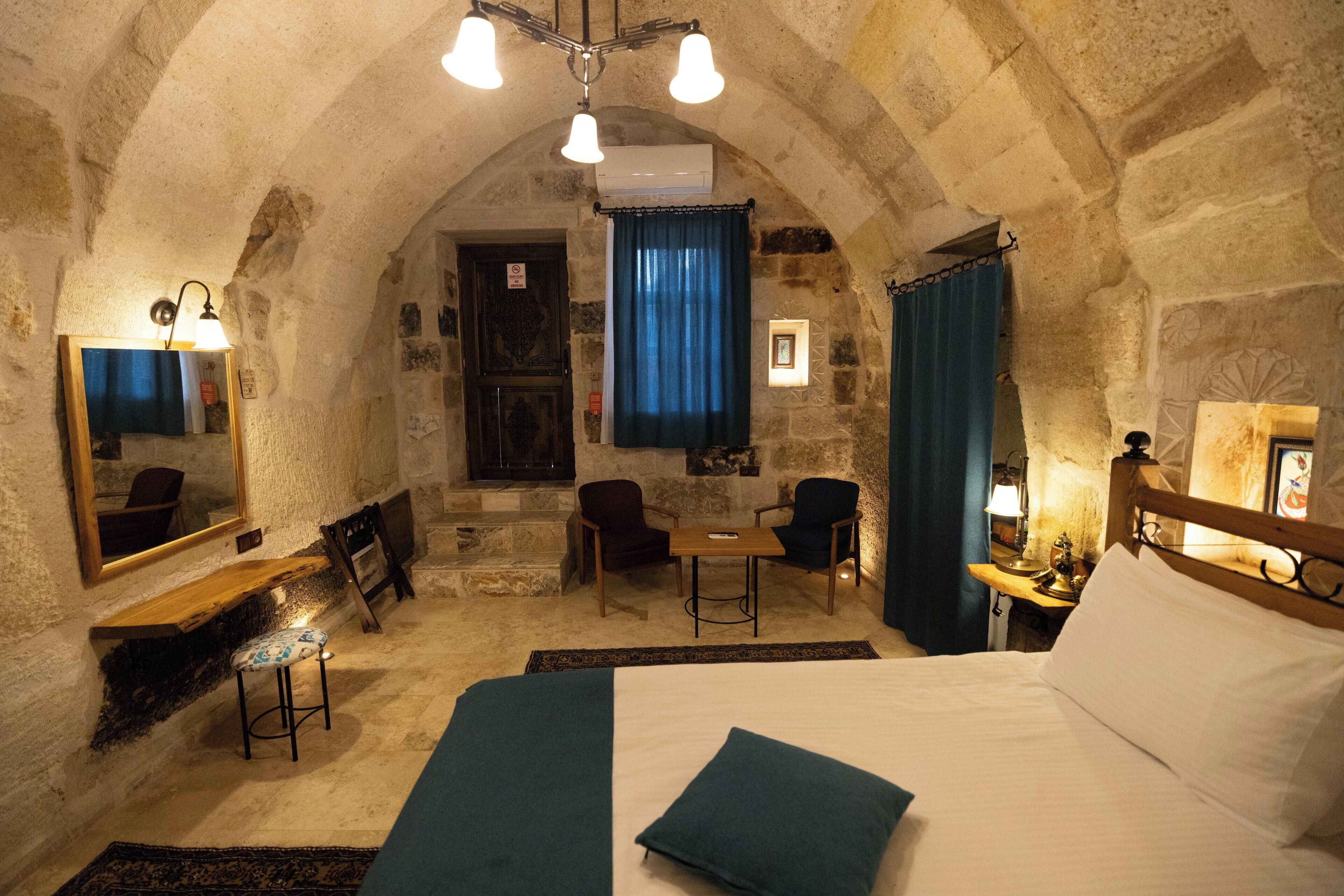 Um quarto acolhedor, com paredes de pedra, uma cama, uma escrivaninha, duas cadeiras e um espelho.