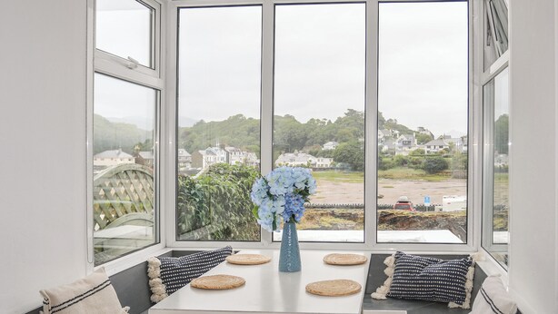 Quarto com mesa e banco brancos, um vaso com flores e vista para casas e vegetação lá fora.