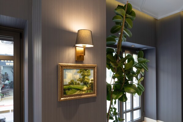 Um candeeiro fixado na parede, um quadro com paisagem enquadrada e uma planta num vaso em um corredor.