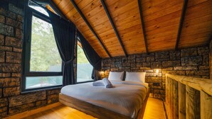 Quarto com teto de madeira, paredes de pedra, uma cama grande e uma janela com cortinas.