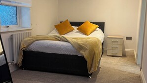 Quarto com cama, duas almofadas, mesa de cabeceira e janela com persianas.