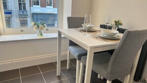 Uma área de refeições com uma mesa branca posta para dois, um vaso de flores brancas e vista para um edifício de tijolos pela janela.