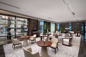 Um hall de hotel moderno com uma grande parede de vidro, diversas áreas de estar e um balcão de recepção.