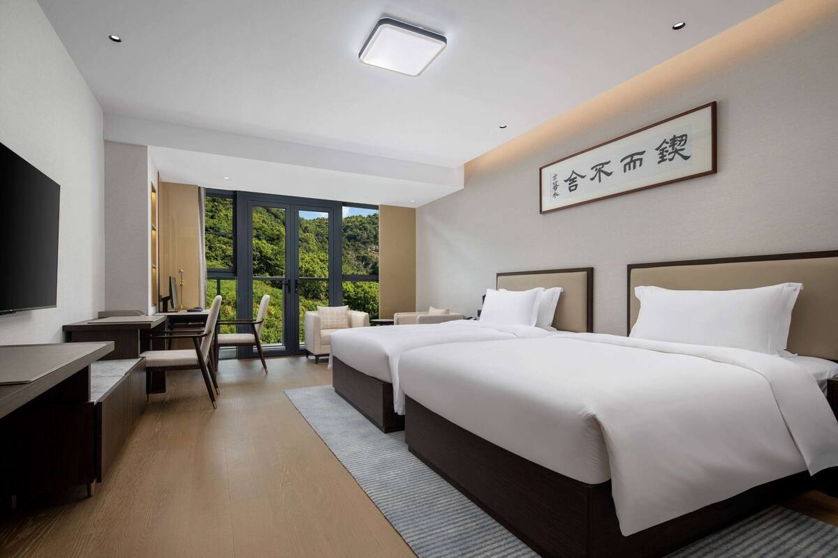 Quarto de hotel moderno com uma cama grande, uma mesa e uma TV. Há uma janela com vista para áreas verdes.
