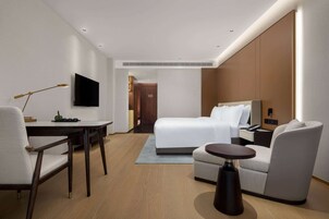 Quarto de hotel moderno com cama, escrivaninha, cadeira, mesinha e televisão.
