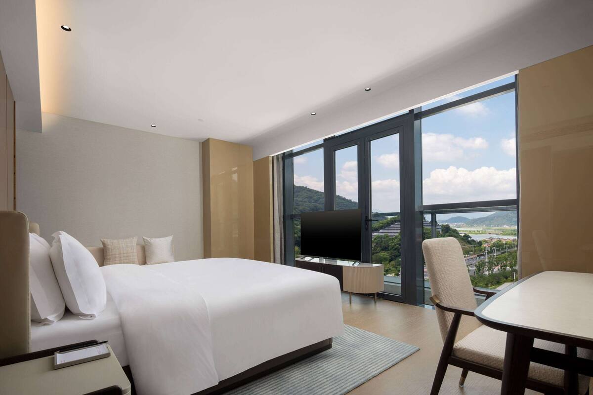 Quarto de hotel moderno com uma cama grande, televisão de tela plana, mesa de jantar e vista para o horizonte urbano pelas amplas janelas.